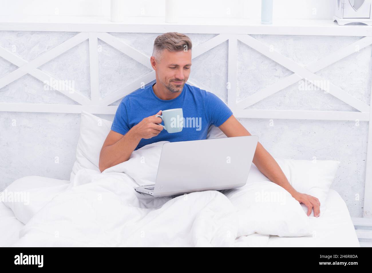 Awake from sleep. Awake man in bed. Happy guy drink tea using laptop ...