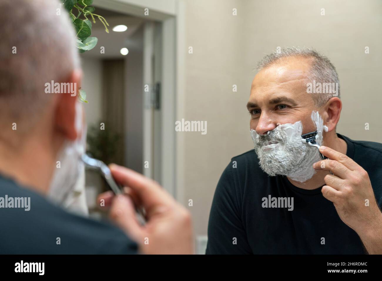 Handsome white-haired beared man shaving off his beard looking in a ...