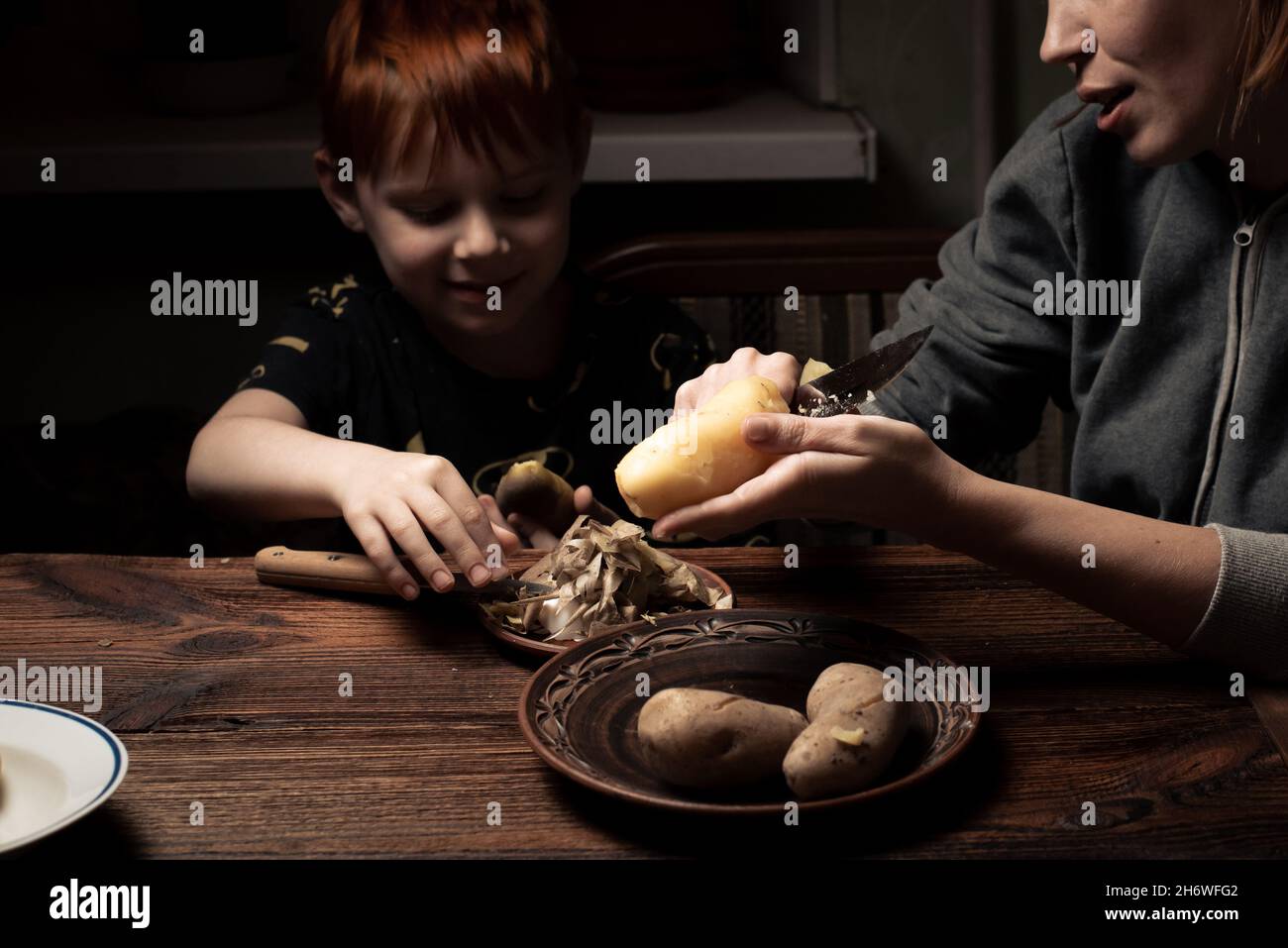 Child with mom peels potatoes on a dark background. A woman teaches her ...