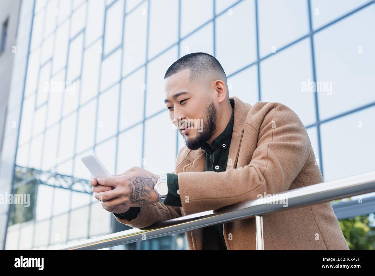 tattooed asian man in beige coat messaging on smartphone near building ...