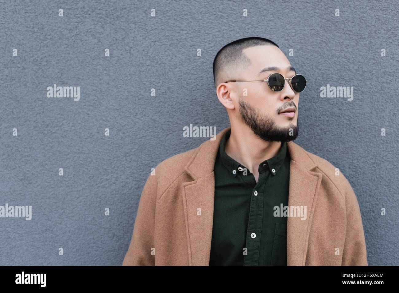asian man in beige coat and sunglasses looking away near grey wall ...