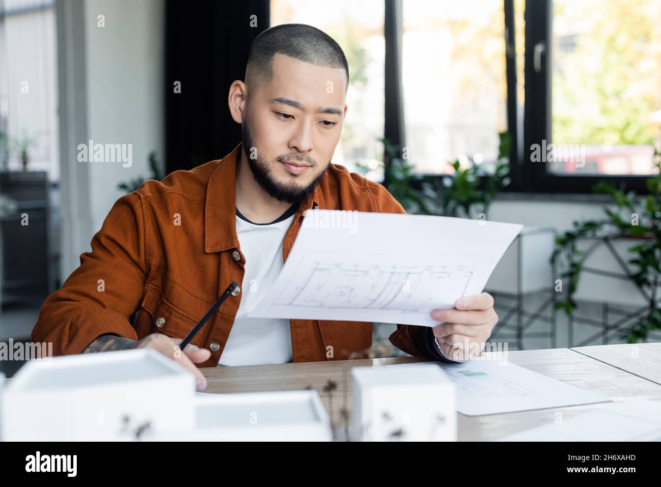 bearded asian architect working with blueprints near blurred house ...
