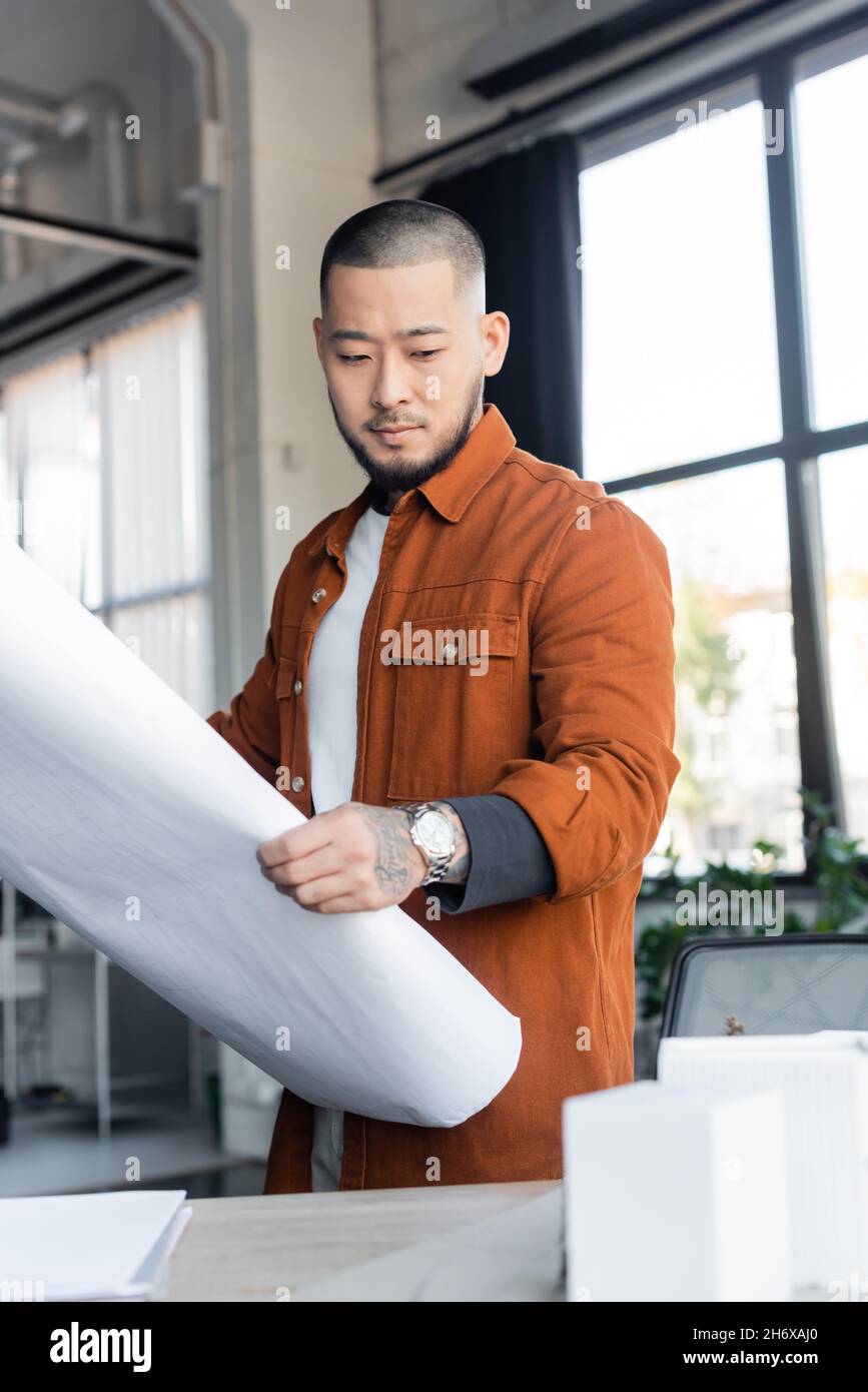 asian architect looking at blurred house models while holding blueprint ...