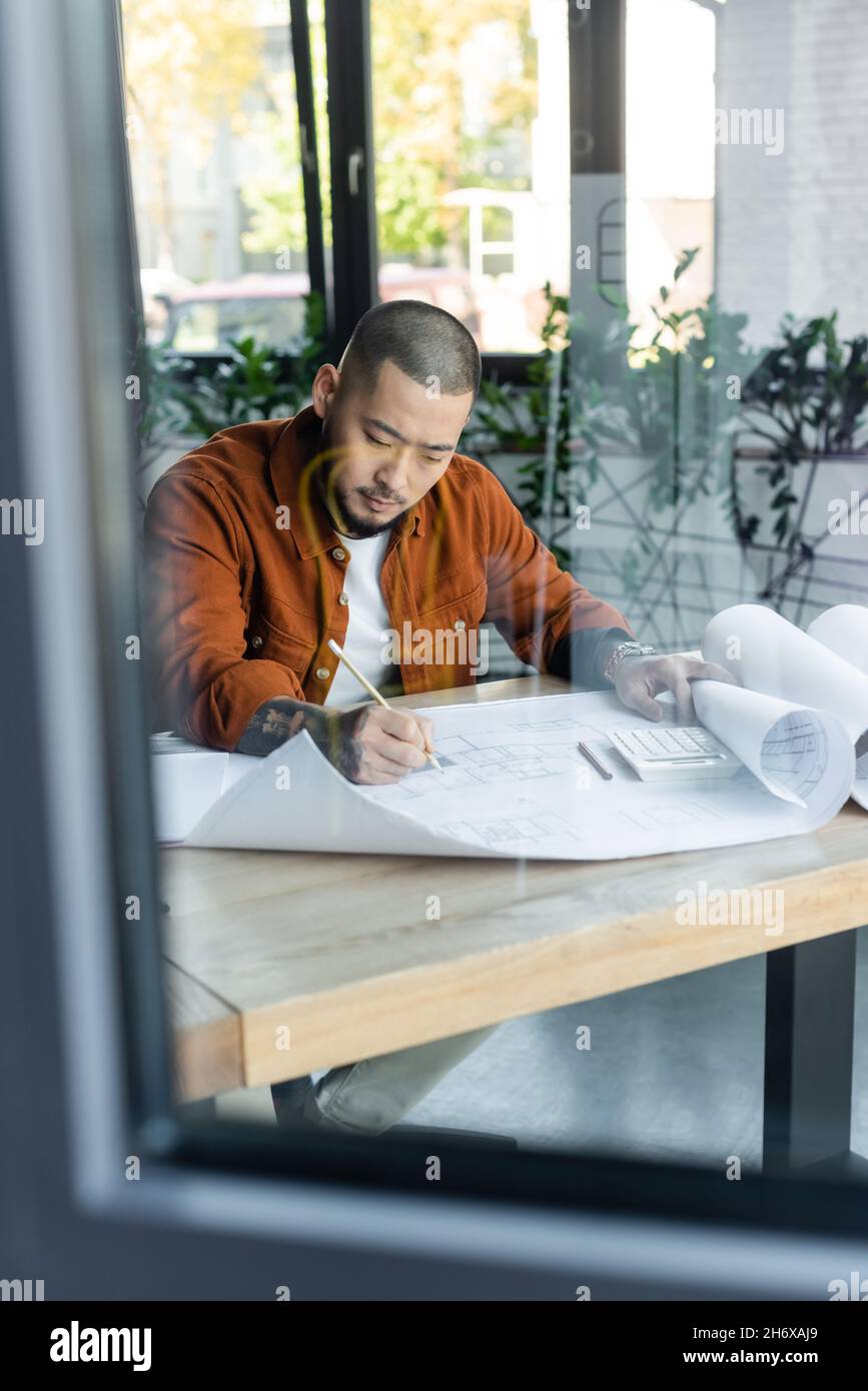 asian architect drawing blueprint at work desk near blurred window ...