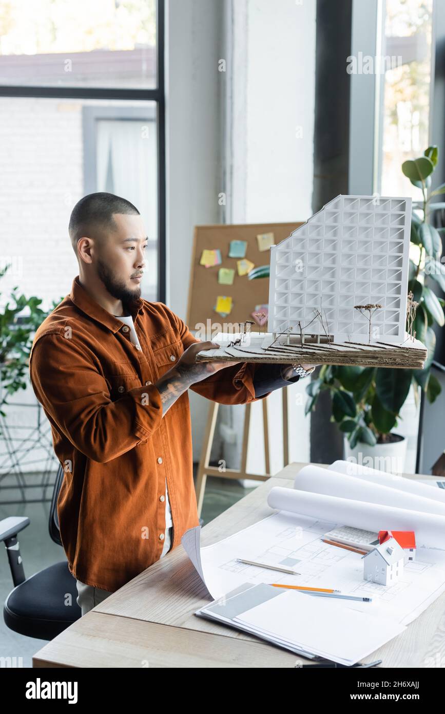 asian architect holding house model near blueprints on desk in office ...