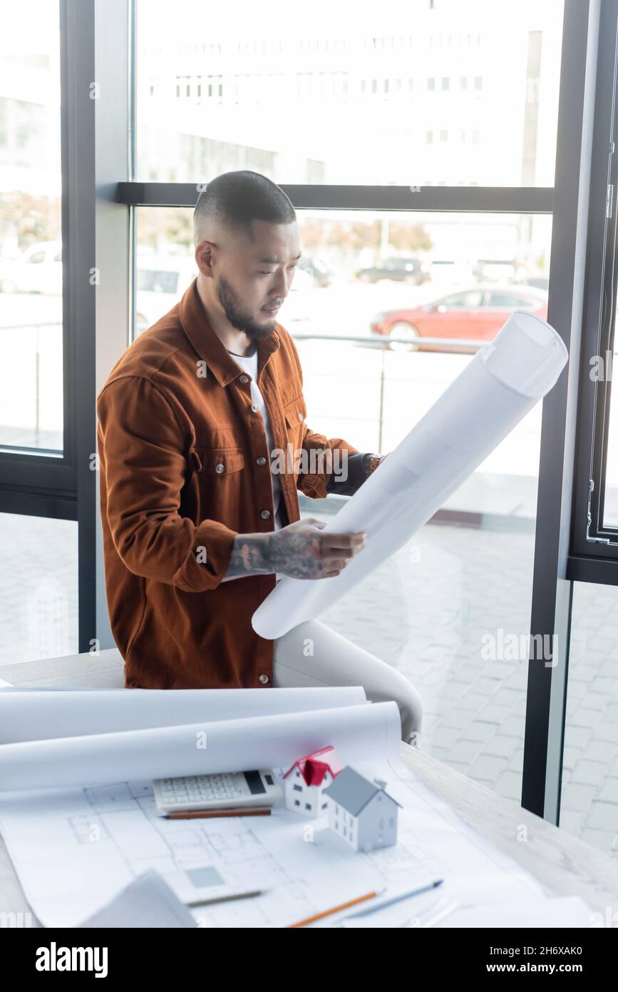 asian architect holding blueprint while sitting on desk near papers and ...