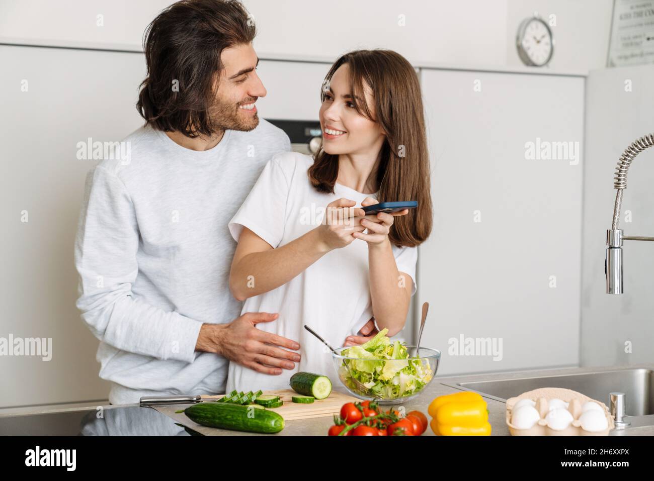 Young multiracial couple using cellphone and hugging while cooking ...