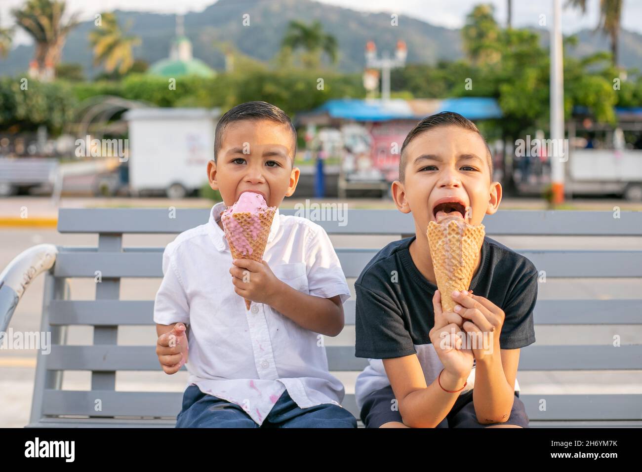 Hispanic cheerful kids sitting on a bench in a park and eating ice ...