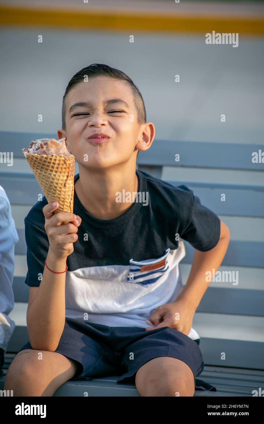 Cheerful Hispanic boy sitting on a bench in a park and eating ice cream ...