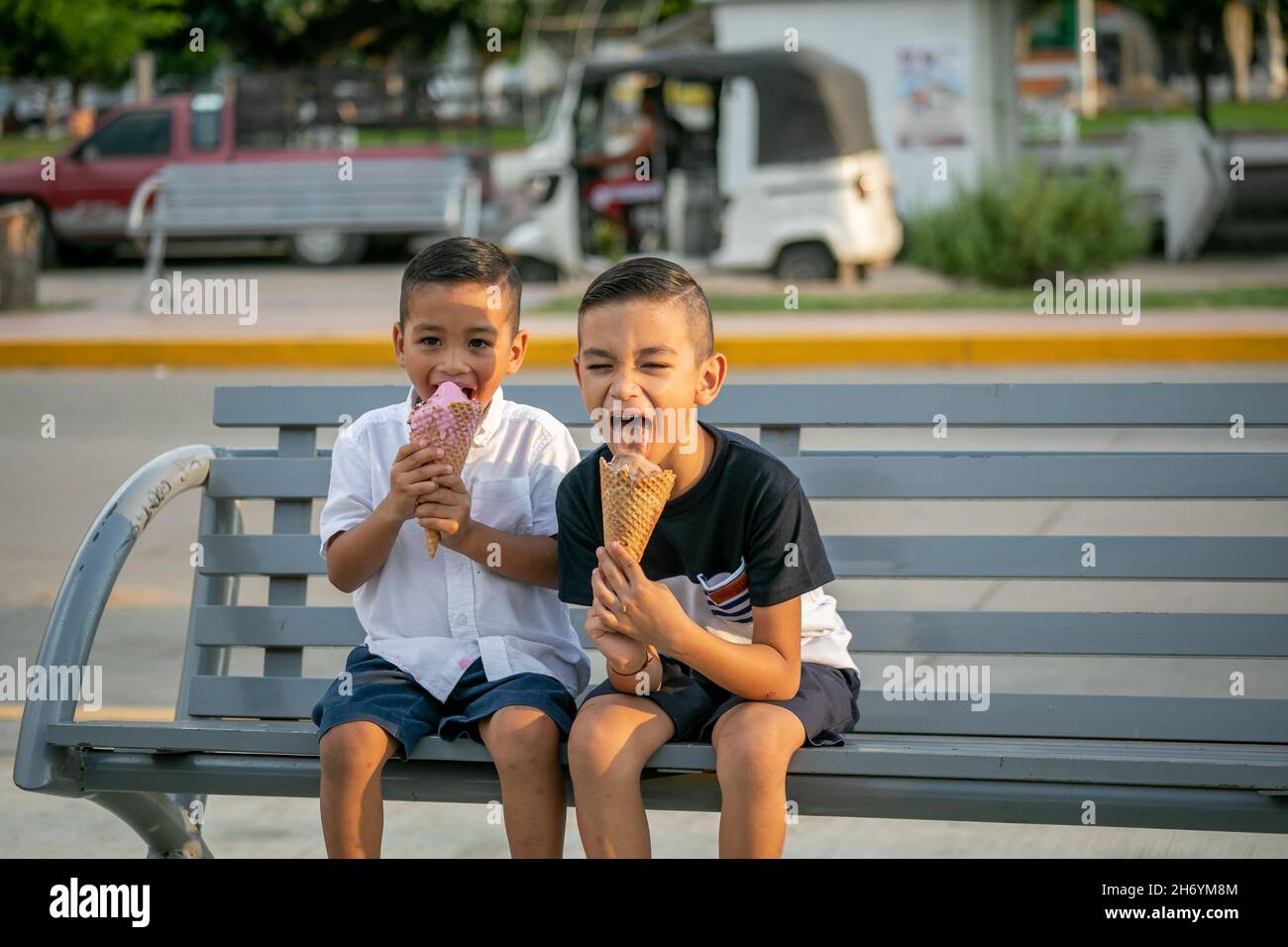 Hispanic cheerful kids sitting on a bench in a park and eating ice ...