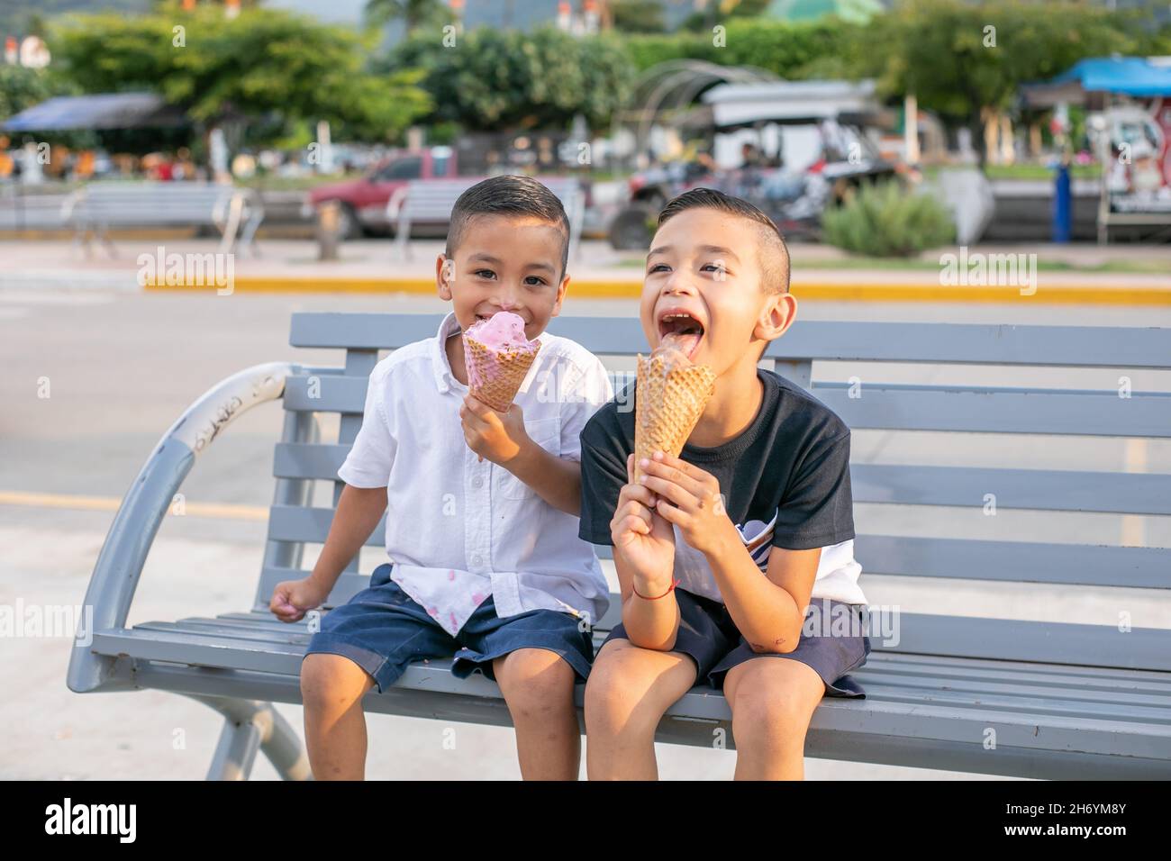 Hispanic cheerful kids sitting on a bench in a park and eating ice ...