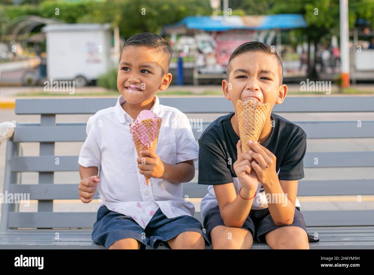 Hispanic cheerful kids sitting on a bench in a park and eating ice ...