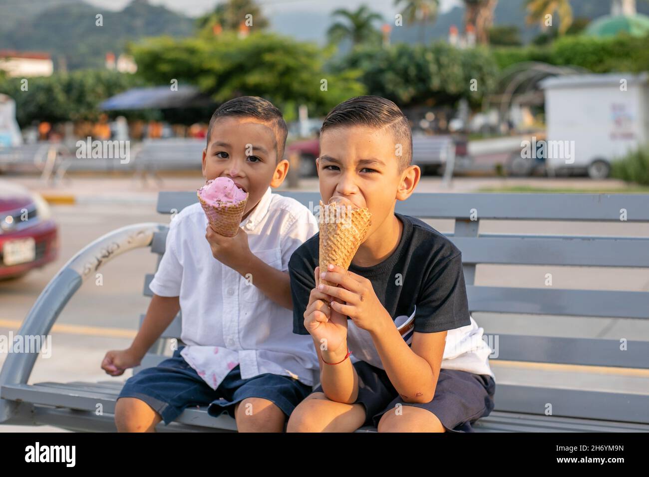 Hispanic cheerful kids sitting on a bench in a park and eating ice ...