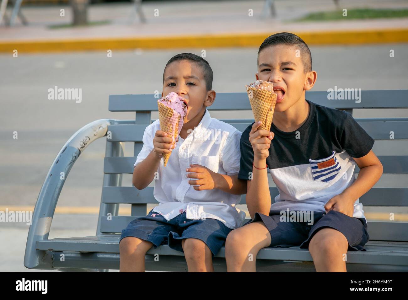 Hispanic cheerful kids sitting on a bench in a park and eating ice ...