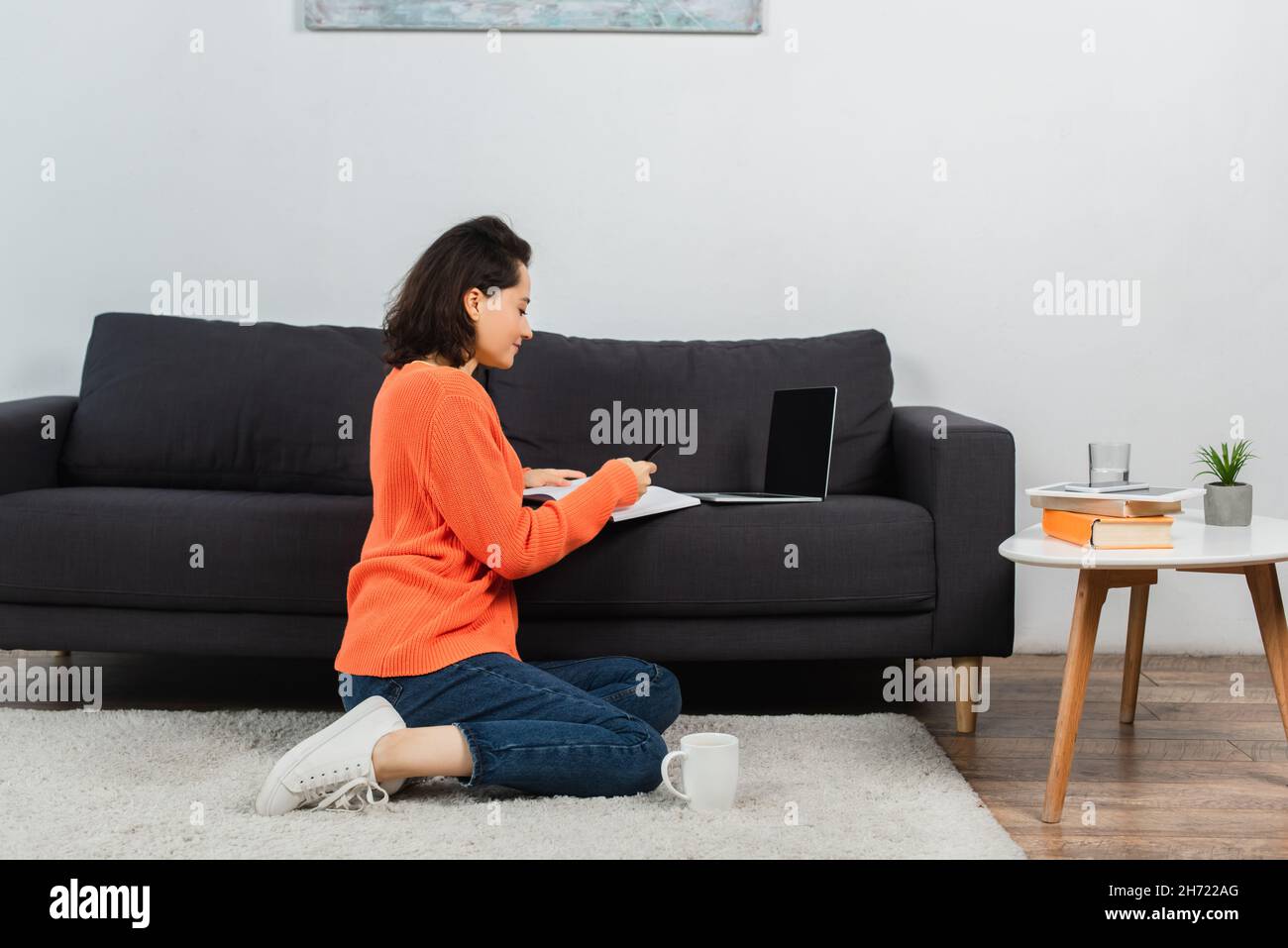woman sitting on carpet and writing in notebook near laptop with blank ...