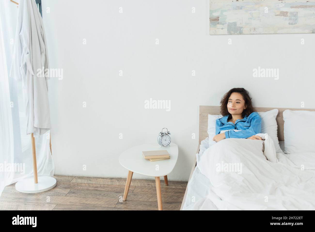 pleased woman lying in bed near bedside table with books and alarm ...