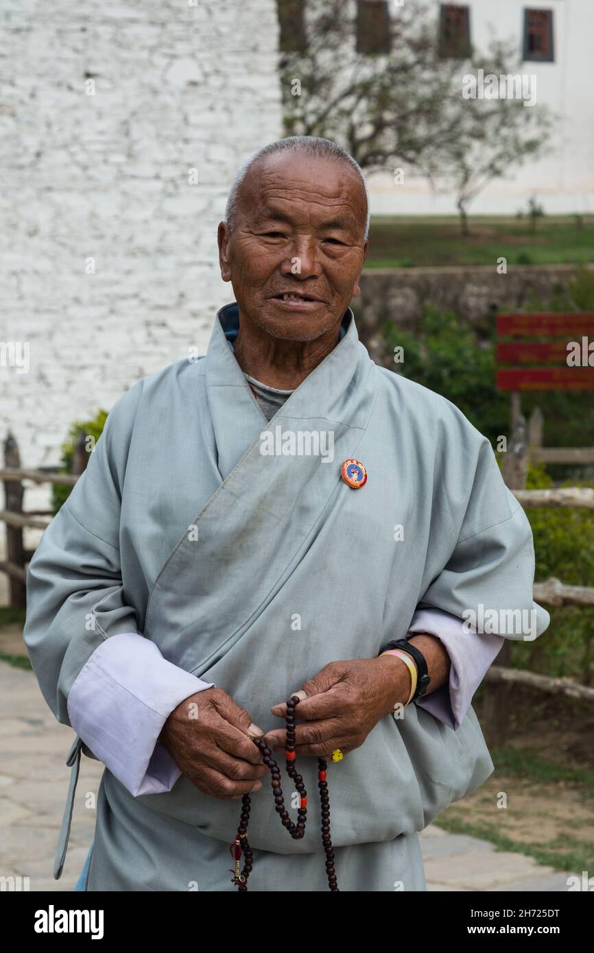 An older Bhutanese man in a traditional gho robe stands with his rosary ...