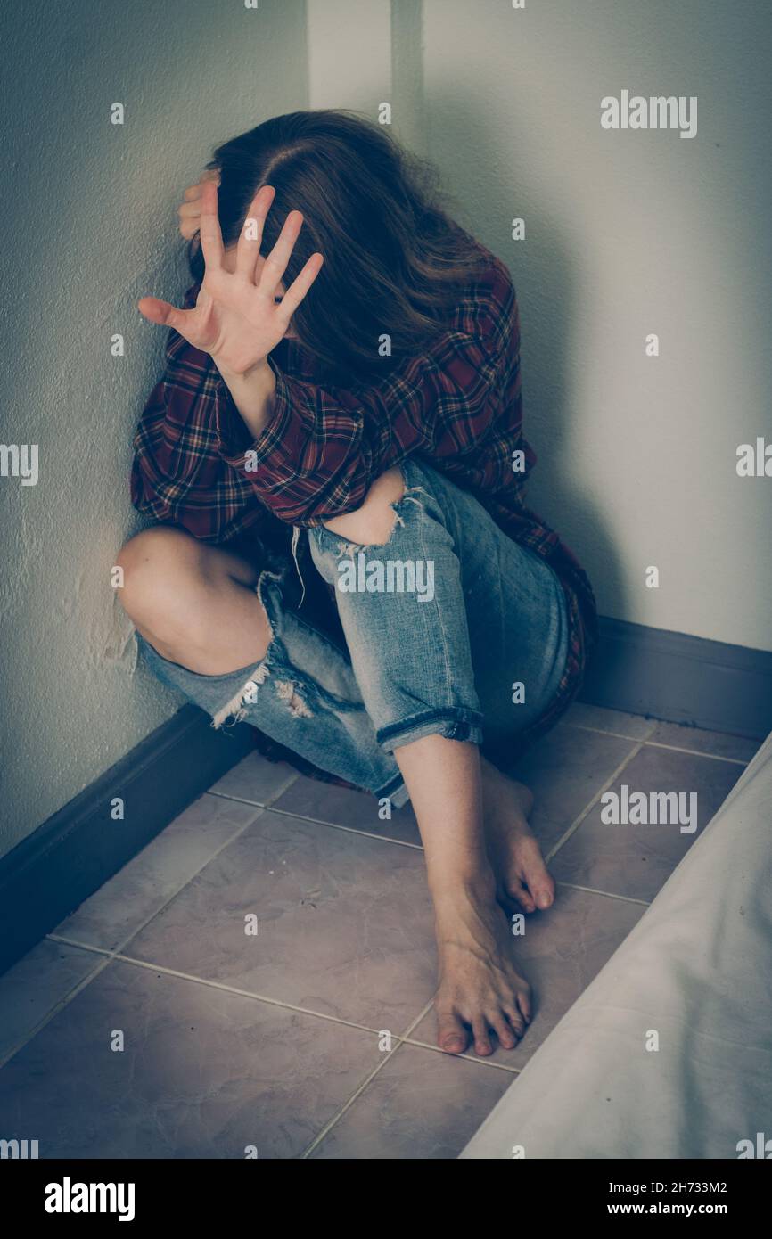 A woman in fear sits on the floor against the wall and covers her face ...
