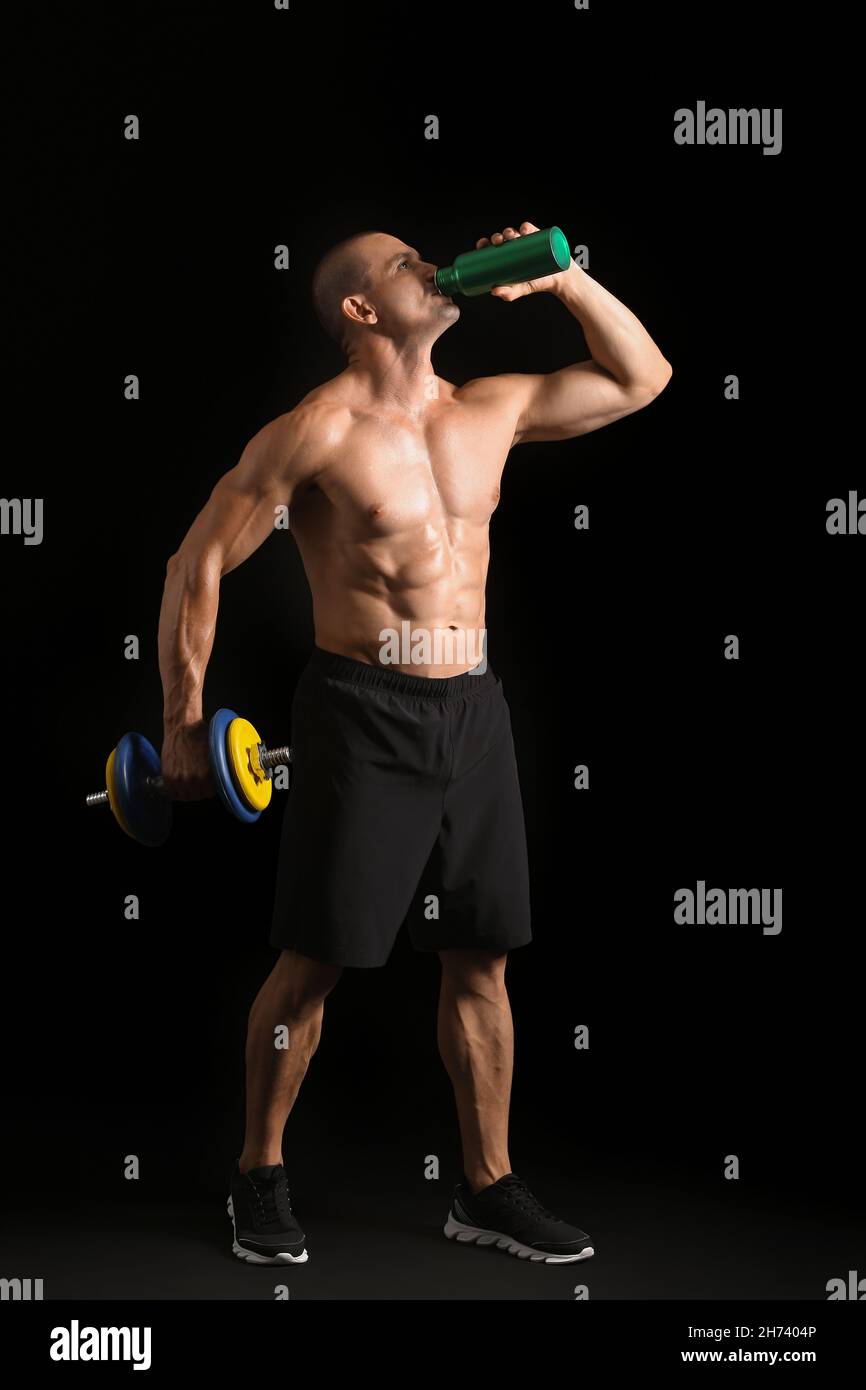 Male bodybuilder with dumbbell drinking protein shake on dark ...