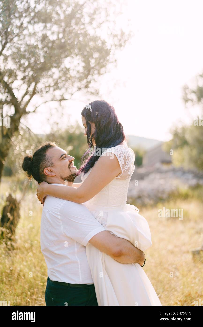 Groom holds bride in his arms against the background of trees on the ...