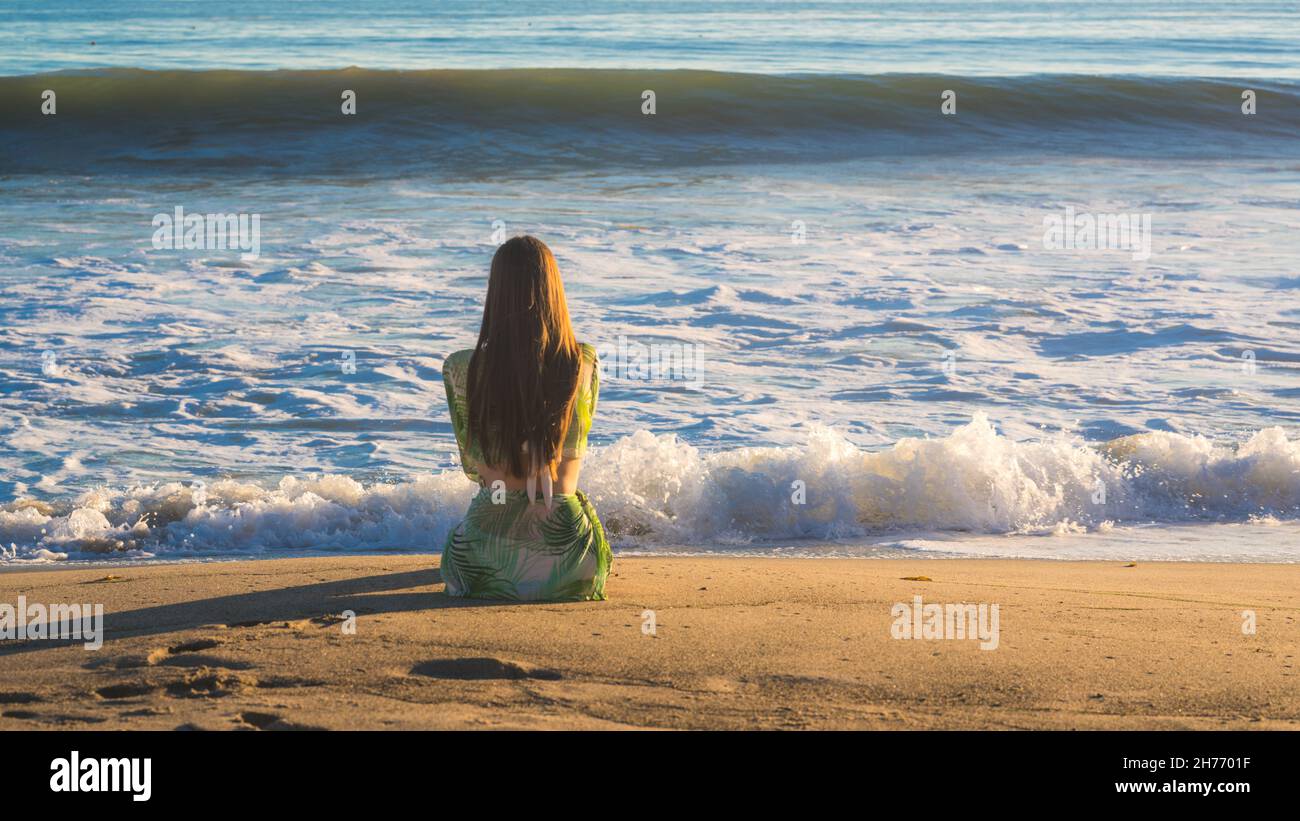 Attractive young woman sitting on the beach looking at the ocean ...