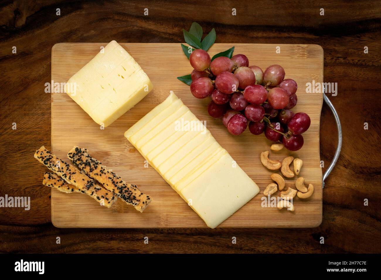 Sliced cheddar cheese block on wood board with grapes, cashew nuts ...