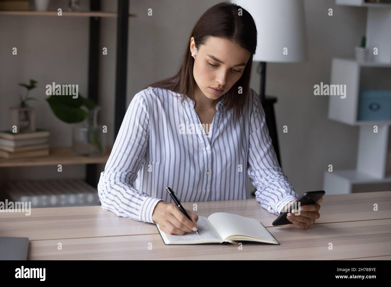 Concentrated young woman writing notes in copybook, using cellphone ...