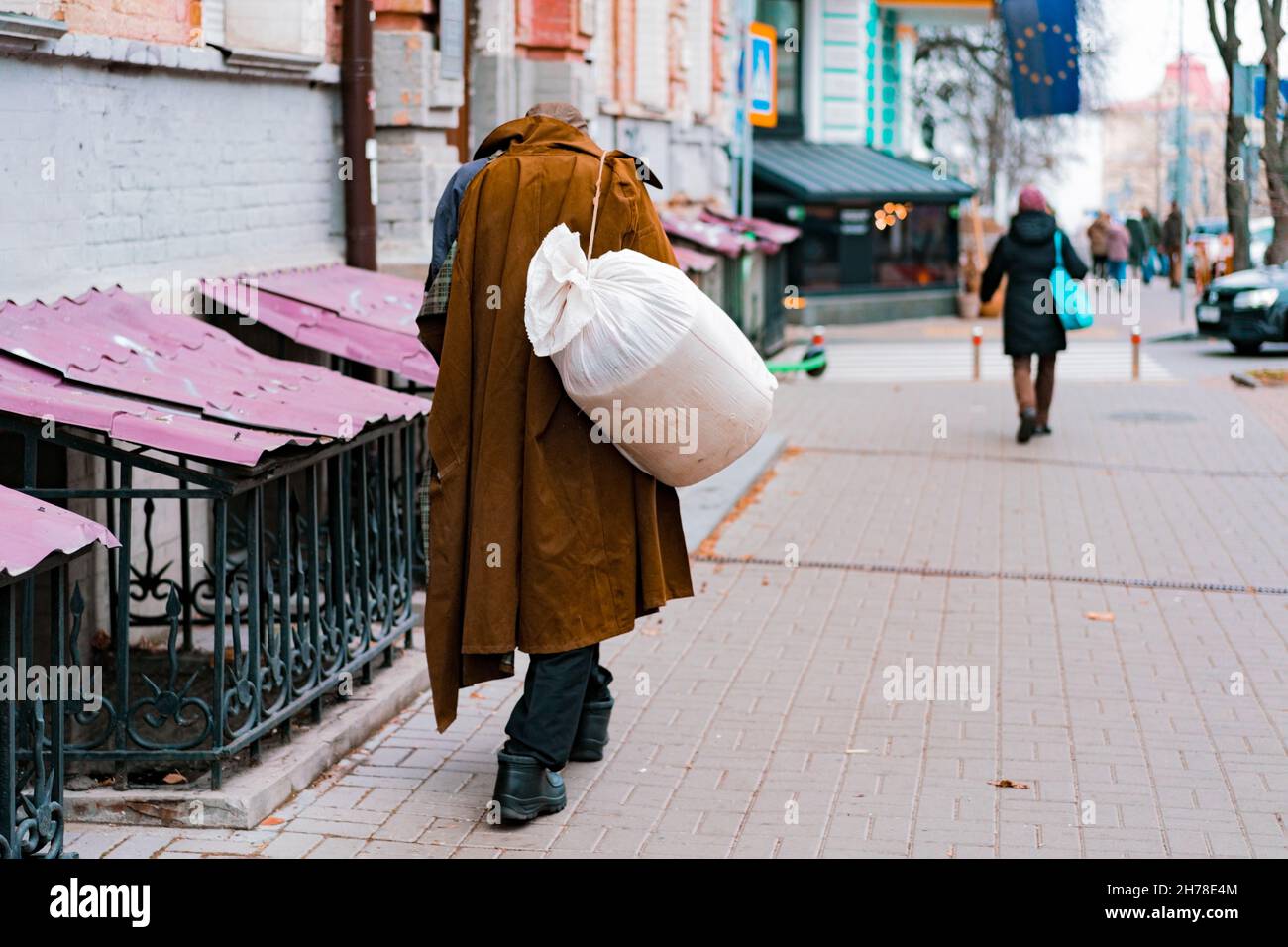 Homeless man in brown long jacket walking city's street with HDPE white ...