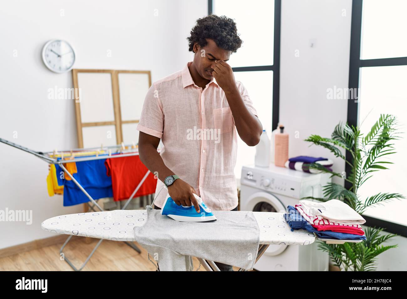 African man with curly hair ironing clothes at home tired rubbing nose ...