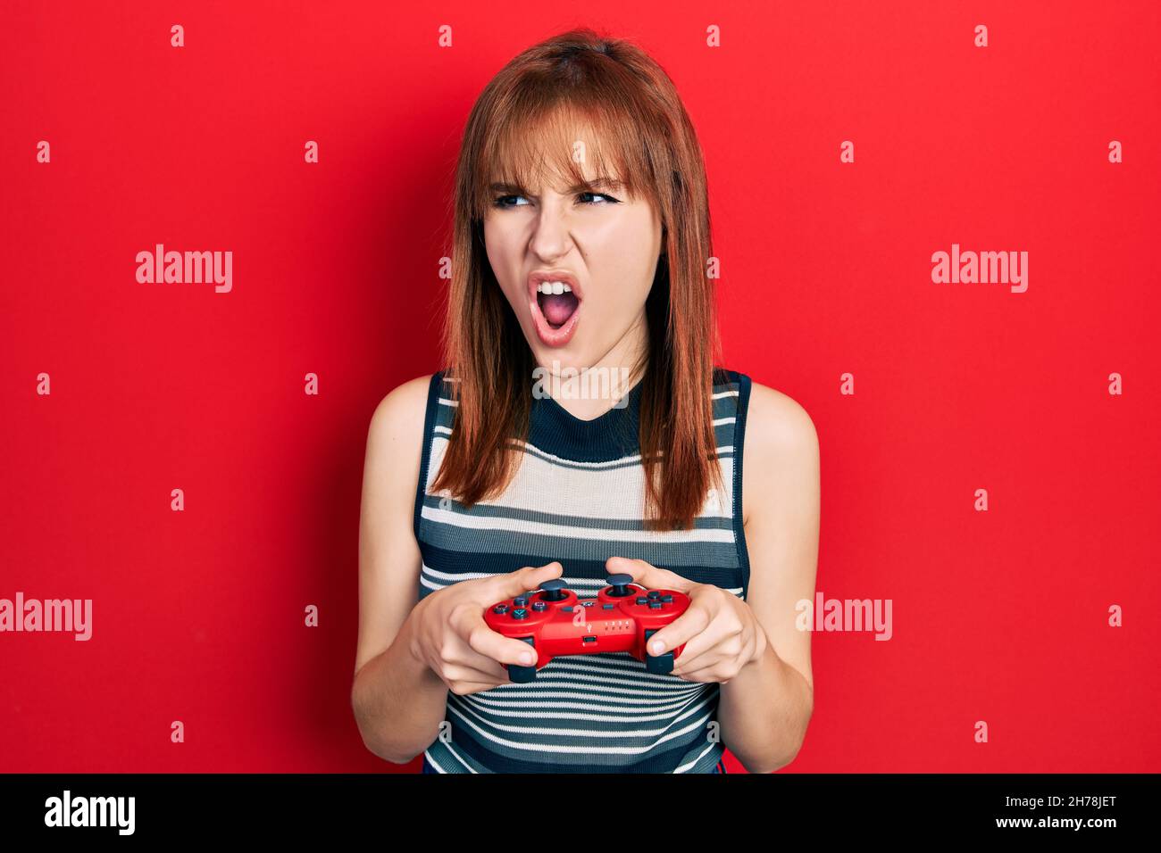 Redhead young woman playing video game holding controller angry and mad ...