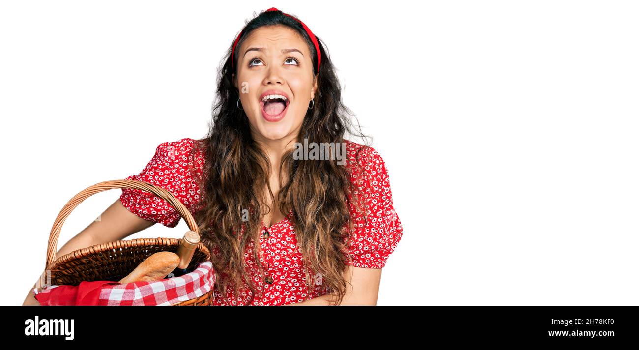Young hispanic girl holding picnic wicker basket with bread angry and ...