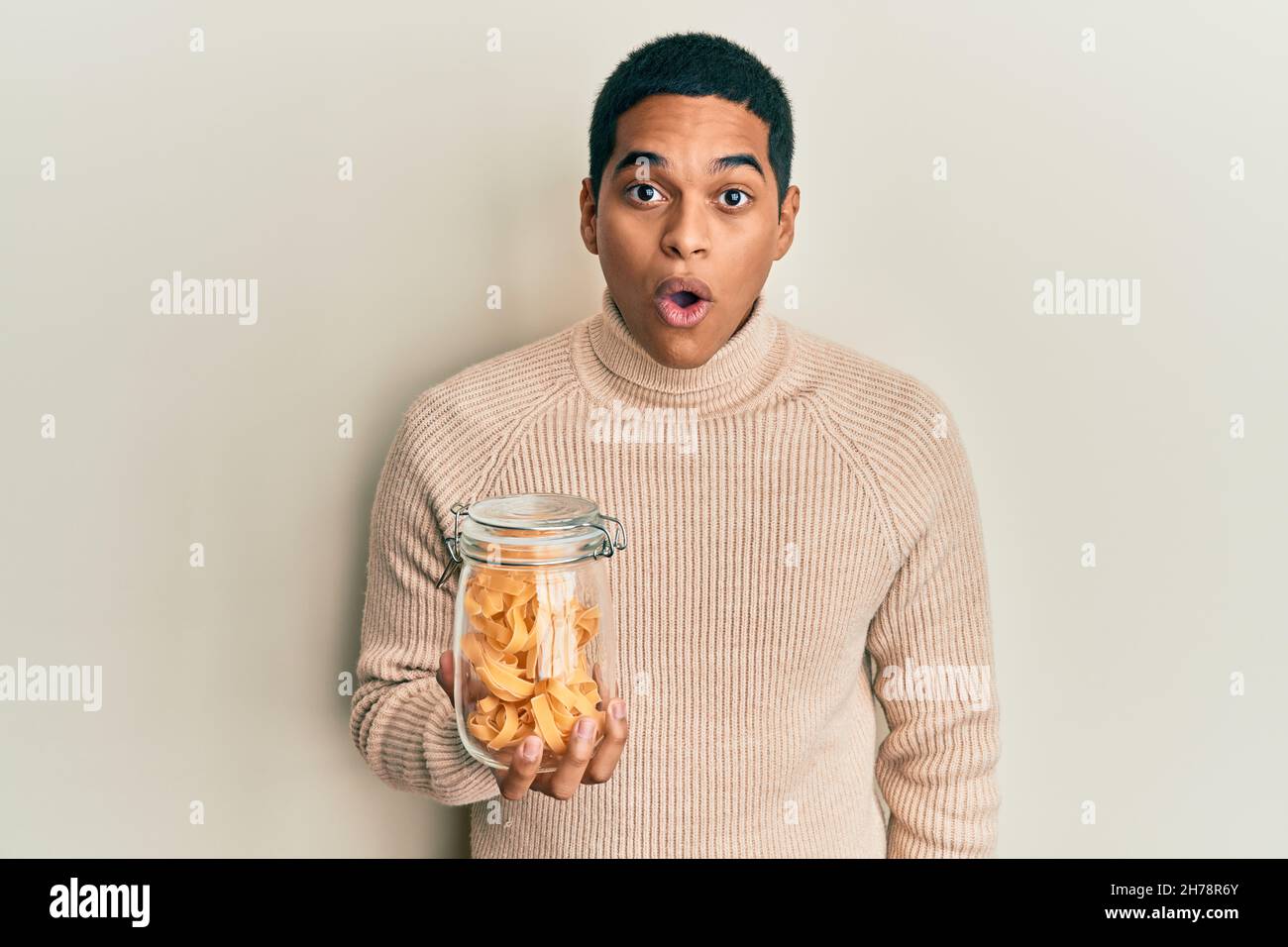 Young handsome hispanic man holding jar with linguine pasta scared and ...
