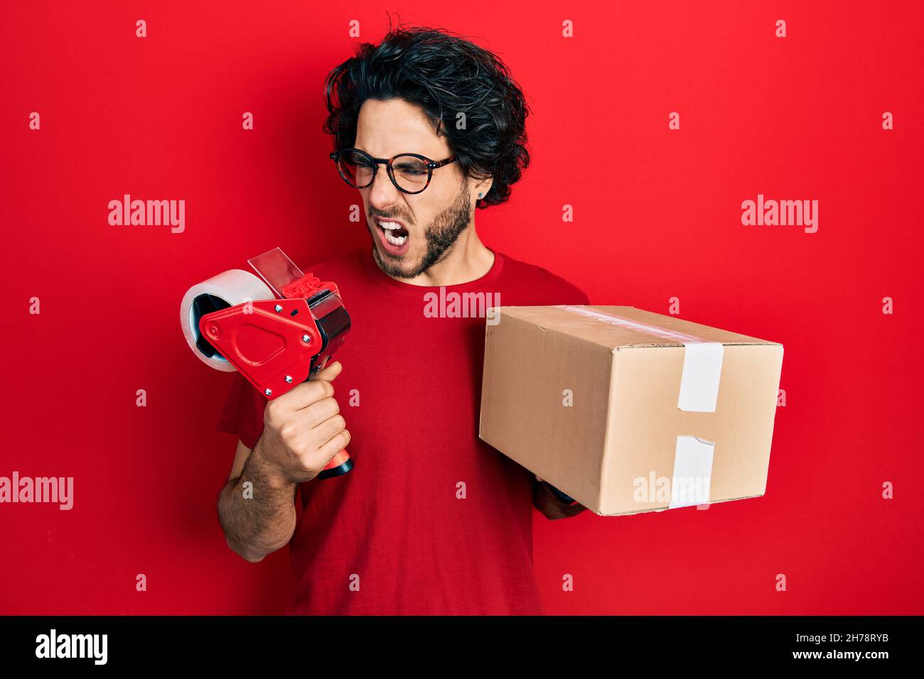 Handsome hispanic man holding packing tape and cardboard box angry and ...