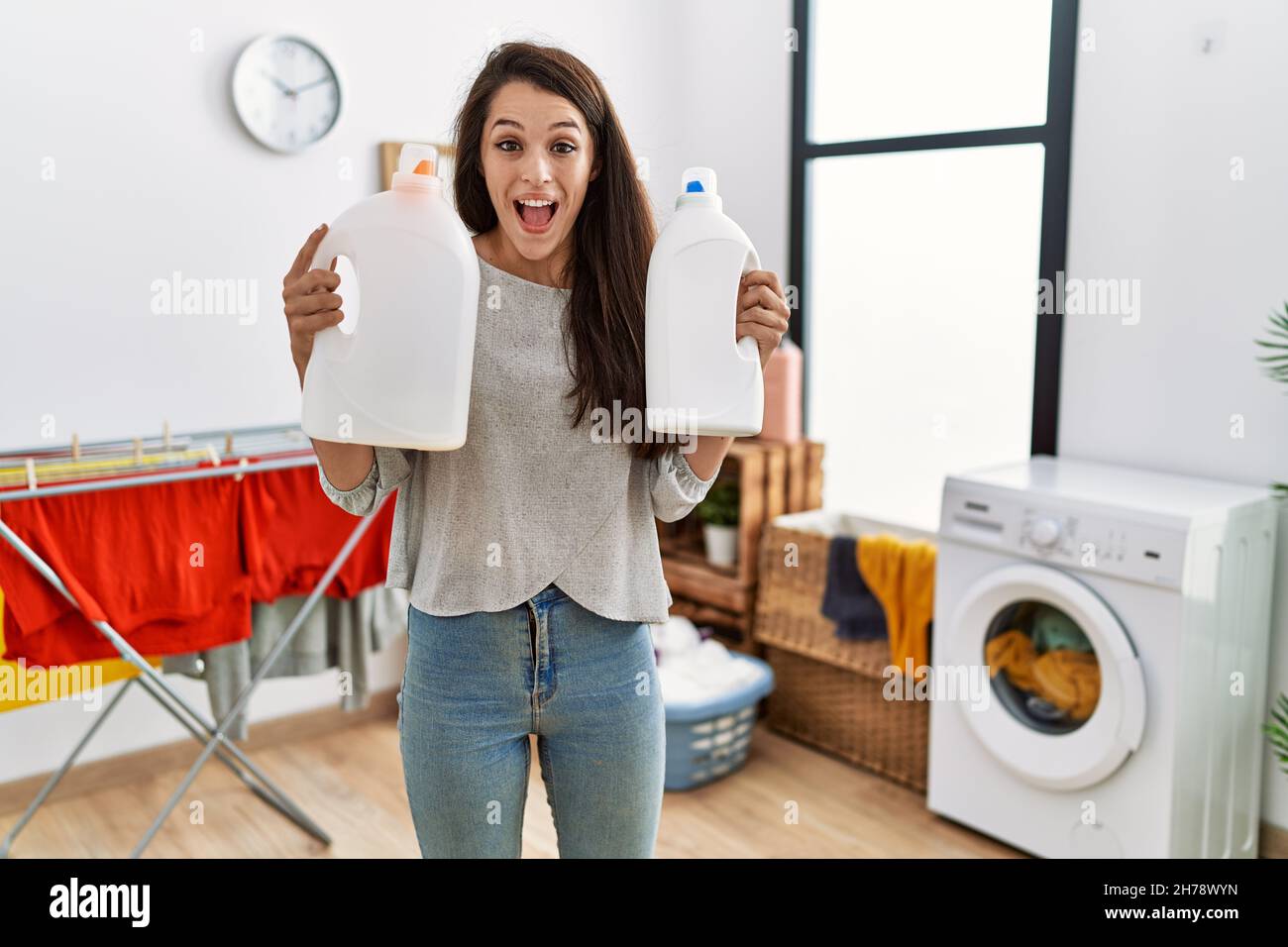 Young brunette woman holding detergent bottle at laundry room celebrating crazy and amazed for ...