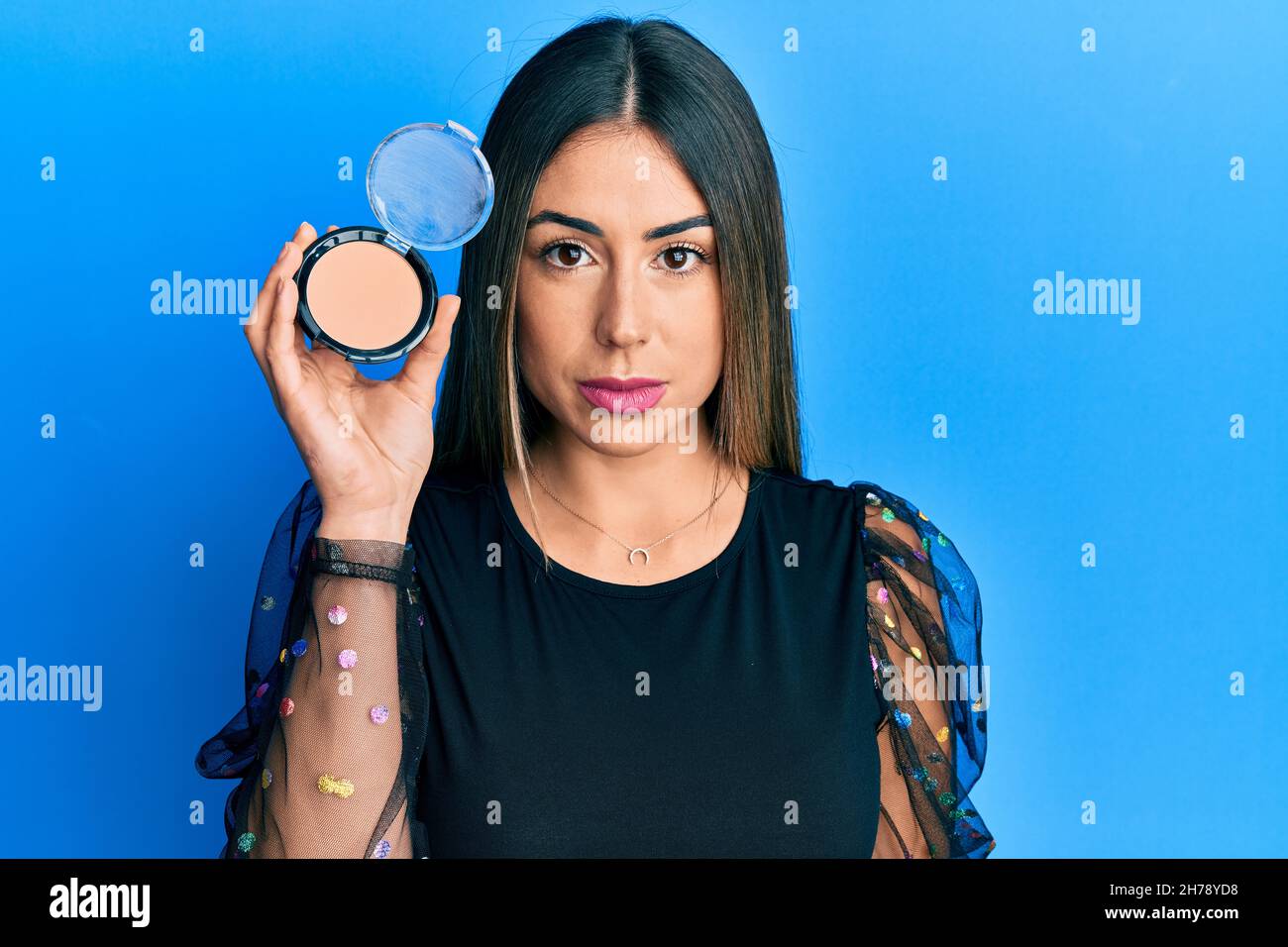 Young hispanic woman holding makeup powder thinking attitude and sober ...