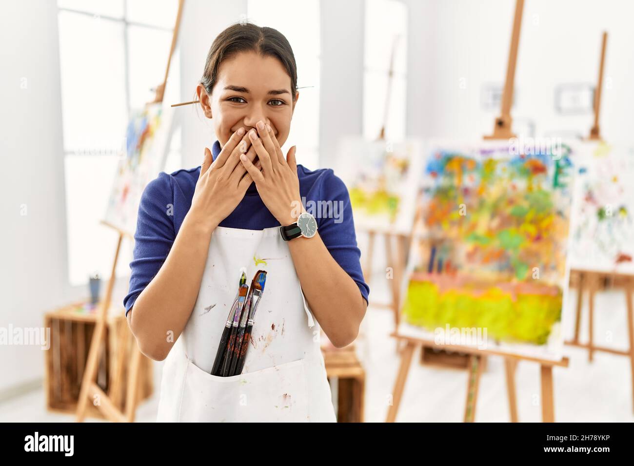 Young brunette woman at art studio laughing and embarrassed giggle covering mouth with hands ...