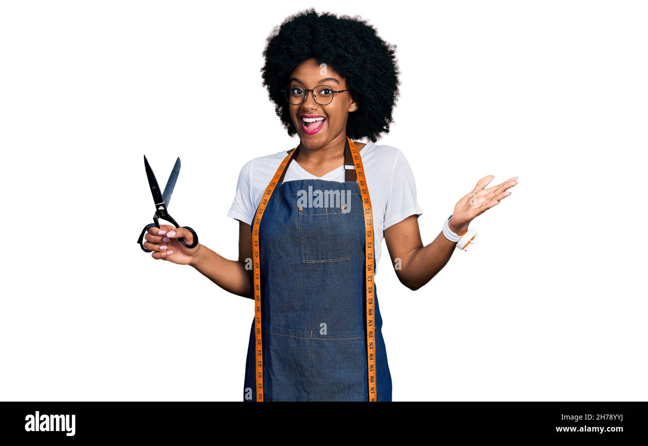 Young african american woman dressmaker designer wearing atelier apron holding scissors ...