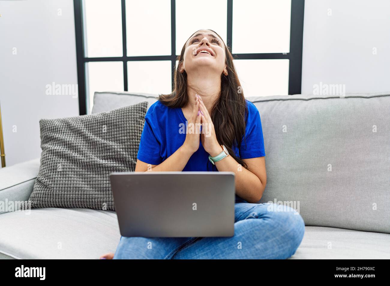 Young brunette woman using laptop at home begging and praying with ...