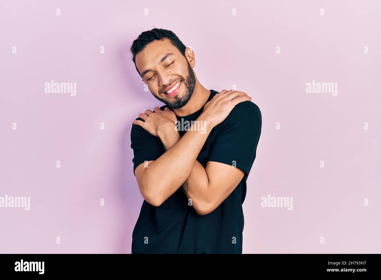 Hispanic man with beard wearing casual black t shirt hugging oneself ...