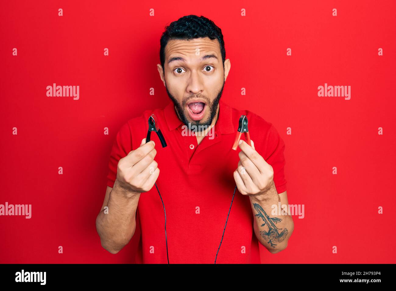 Hispanic man with beard holding battery clamps afraid and shocked with ...