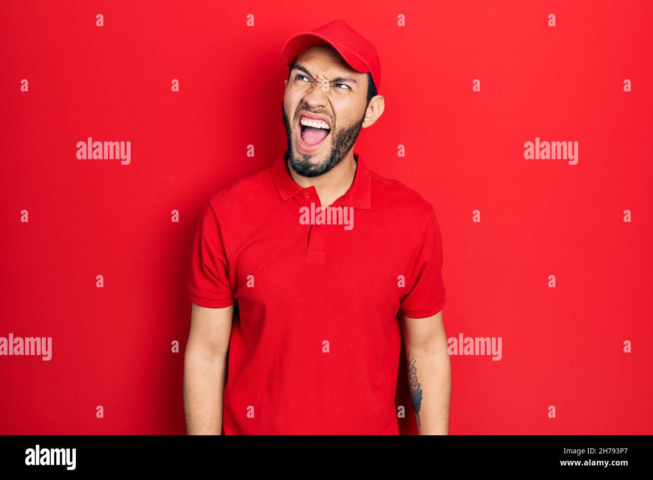 Hispanic man with beard wearing delivery uniform and cap angry and mad ...