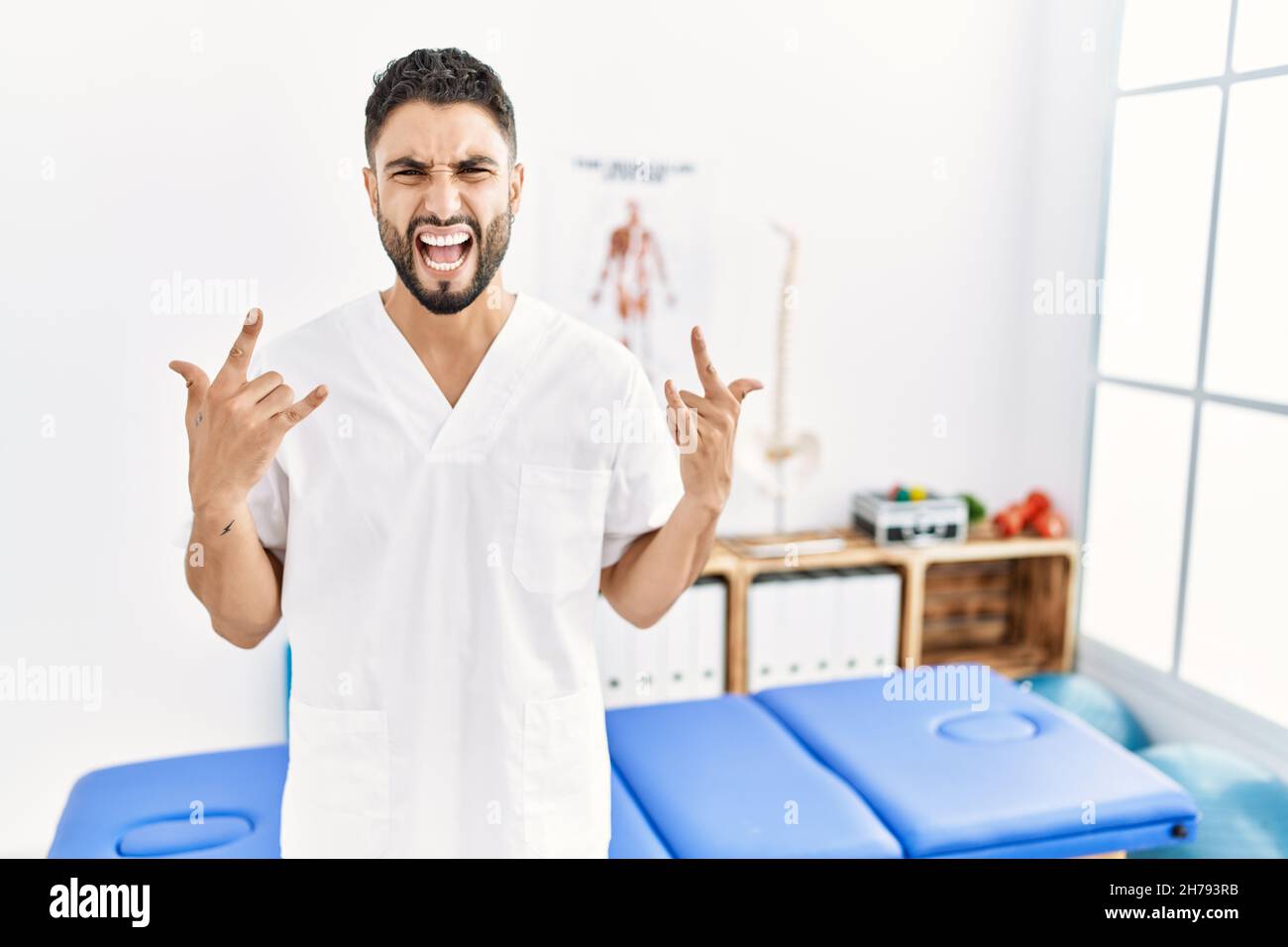 Young handsome man with beard working at pain recovery clinic shouting ...