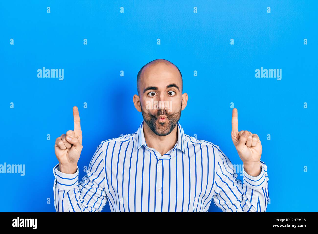 Young bald man pointing up with fingers making fish face with mouth and ...