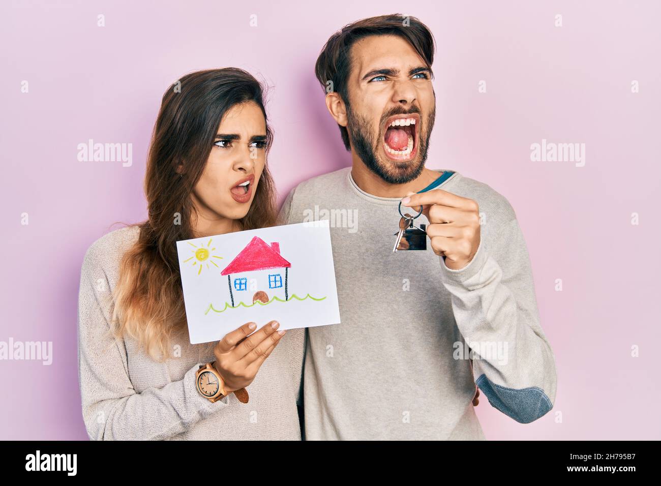 Young hispanic couple holding house draw and key of new home angry and ...