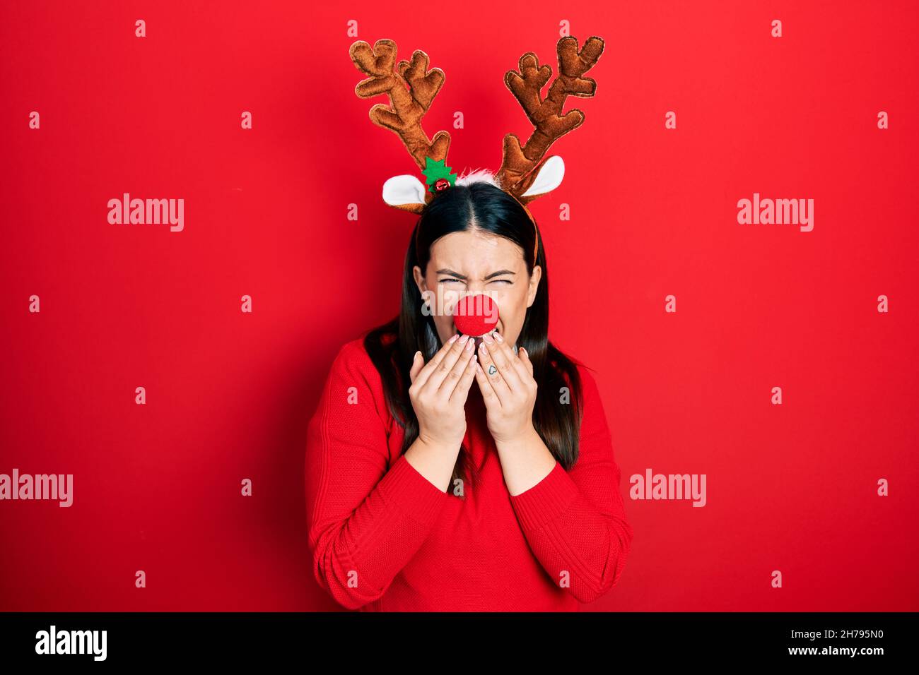 Young hispanic woman wearing deer christmas hat and red nose laughing ...