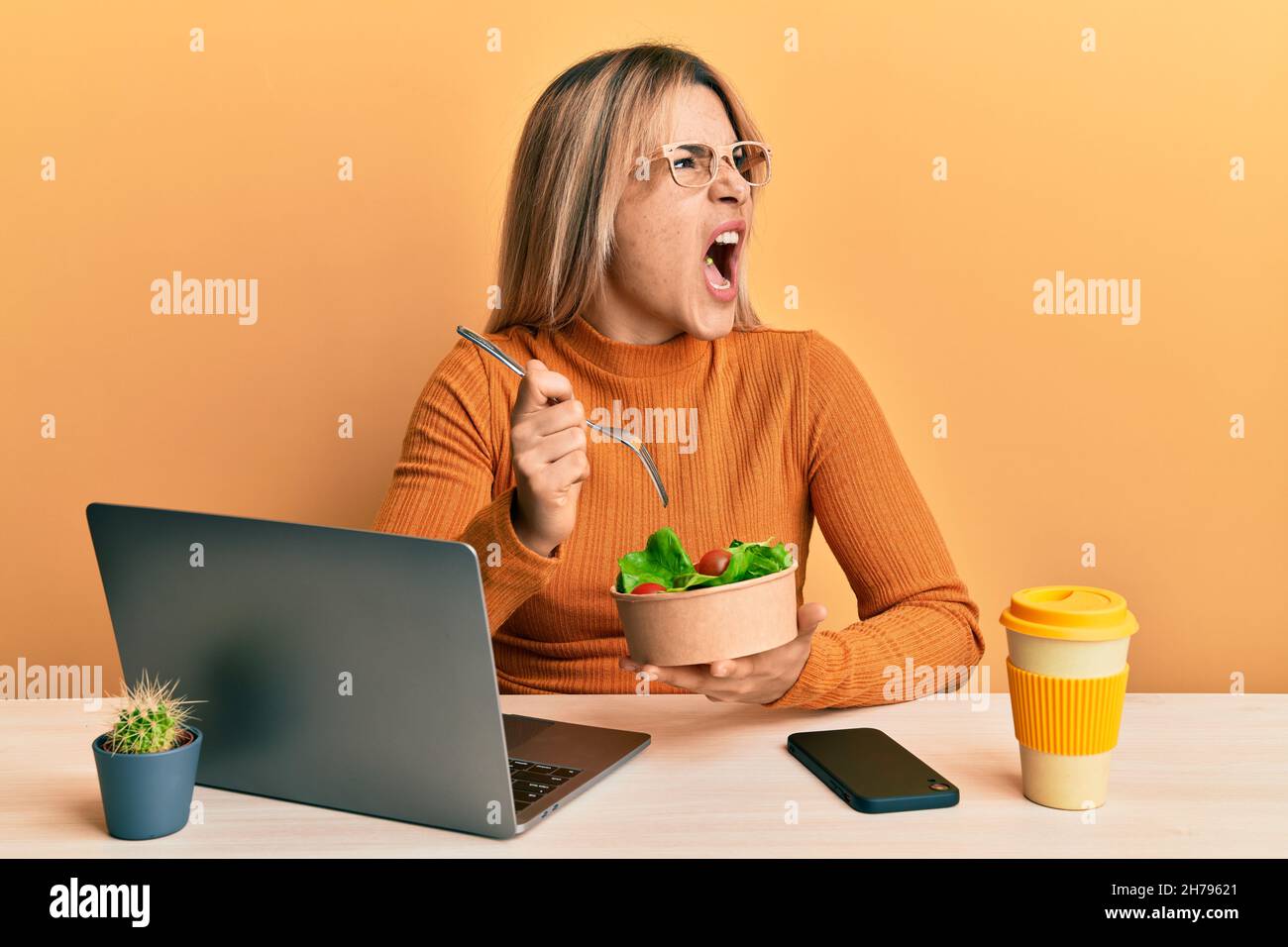 Young caucasian woman working at the office eating healthy salad angry ...