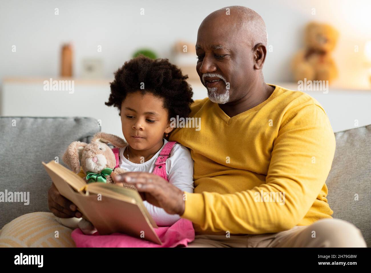 Serious black small girl with toy and elderly man reading book with ...