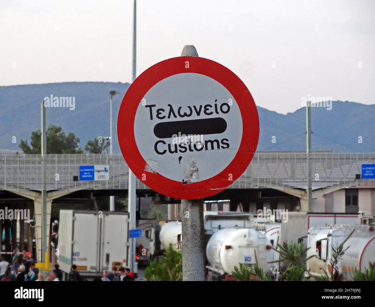 A closeup of a round "customs" Greek road sign surrounded by buildings ...