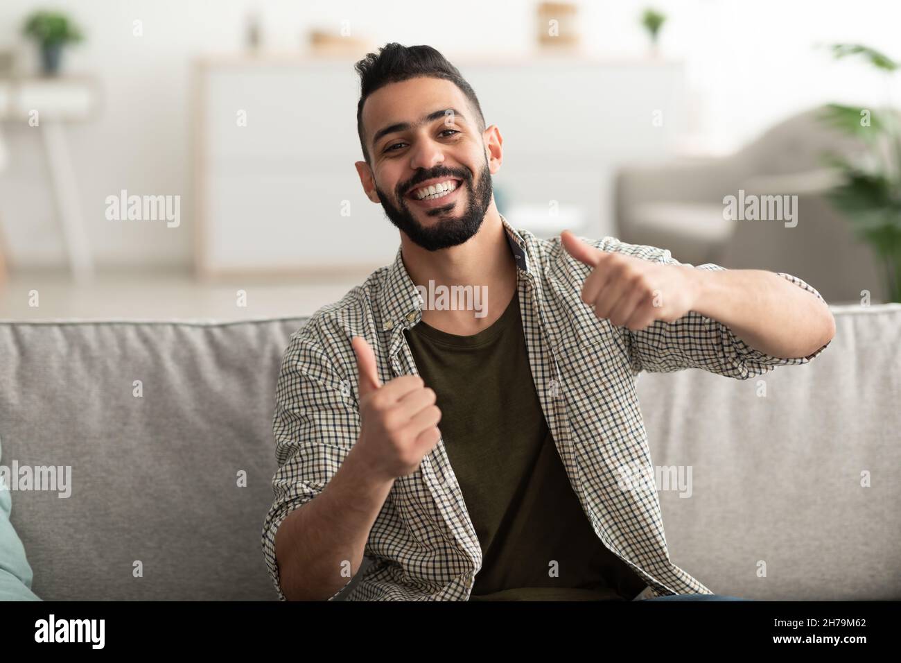 Cheerful young Arab guy showing thumbs up gesture with both hands and smiling at camera, sitting ...
