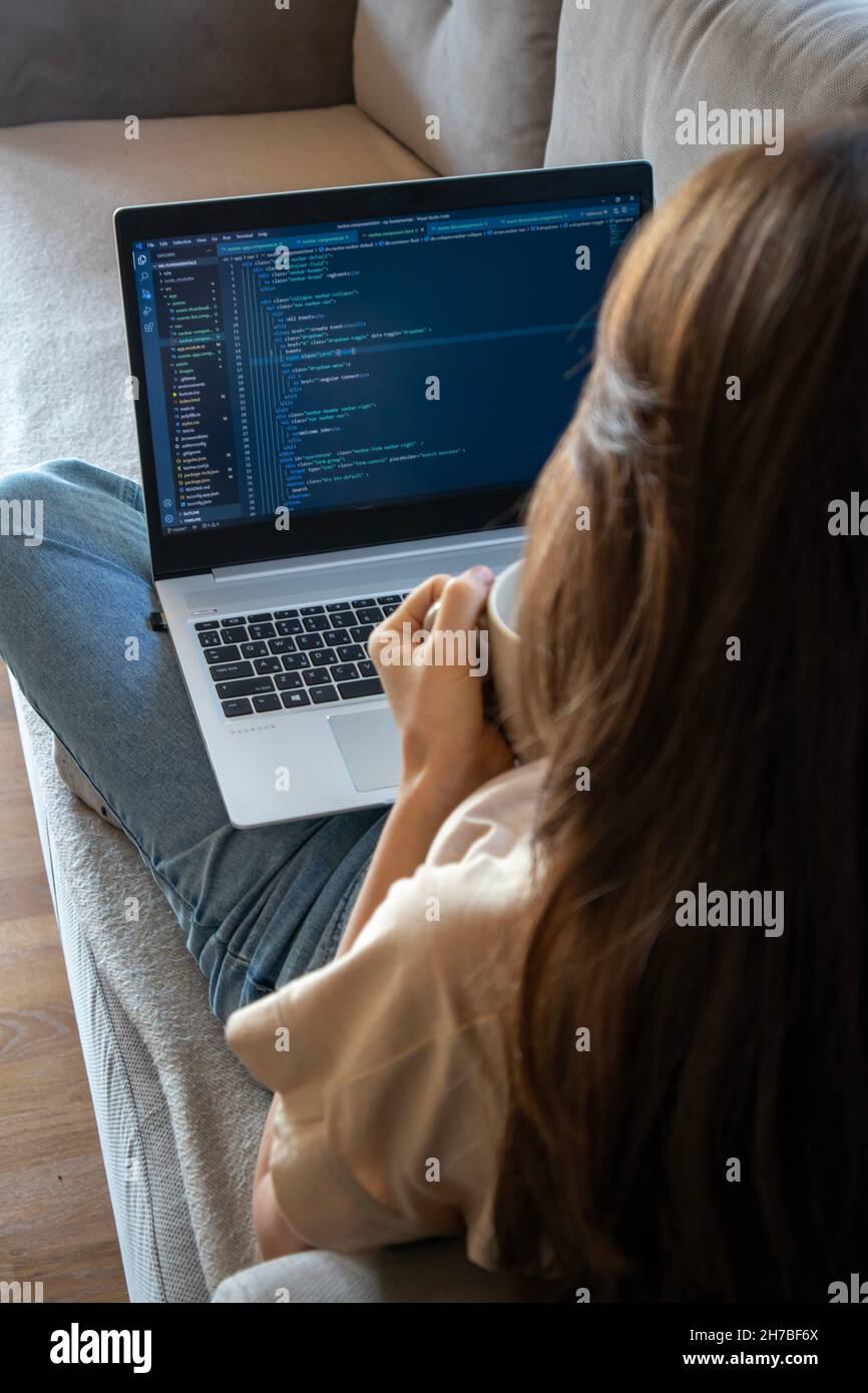 A young woman programmer is coding. A woman is drinking tea while working on a laptop and ...